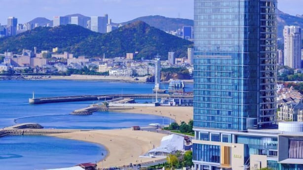 Paisagem costeira com um moderno arranha-céu de vidro em destaque, uma praia e um horizonte montanhoso.