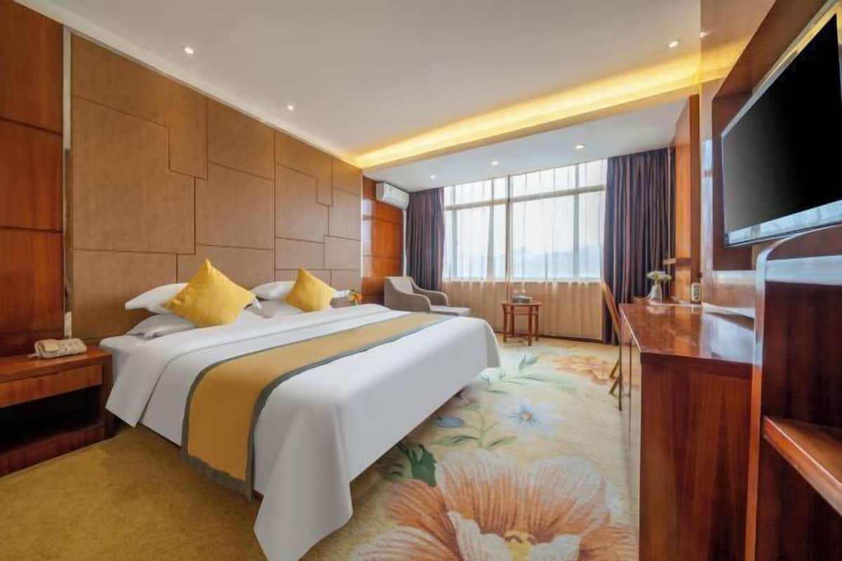 Quarto de hotel com cama, escrivaninha, cadeira, televisão e janela.