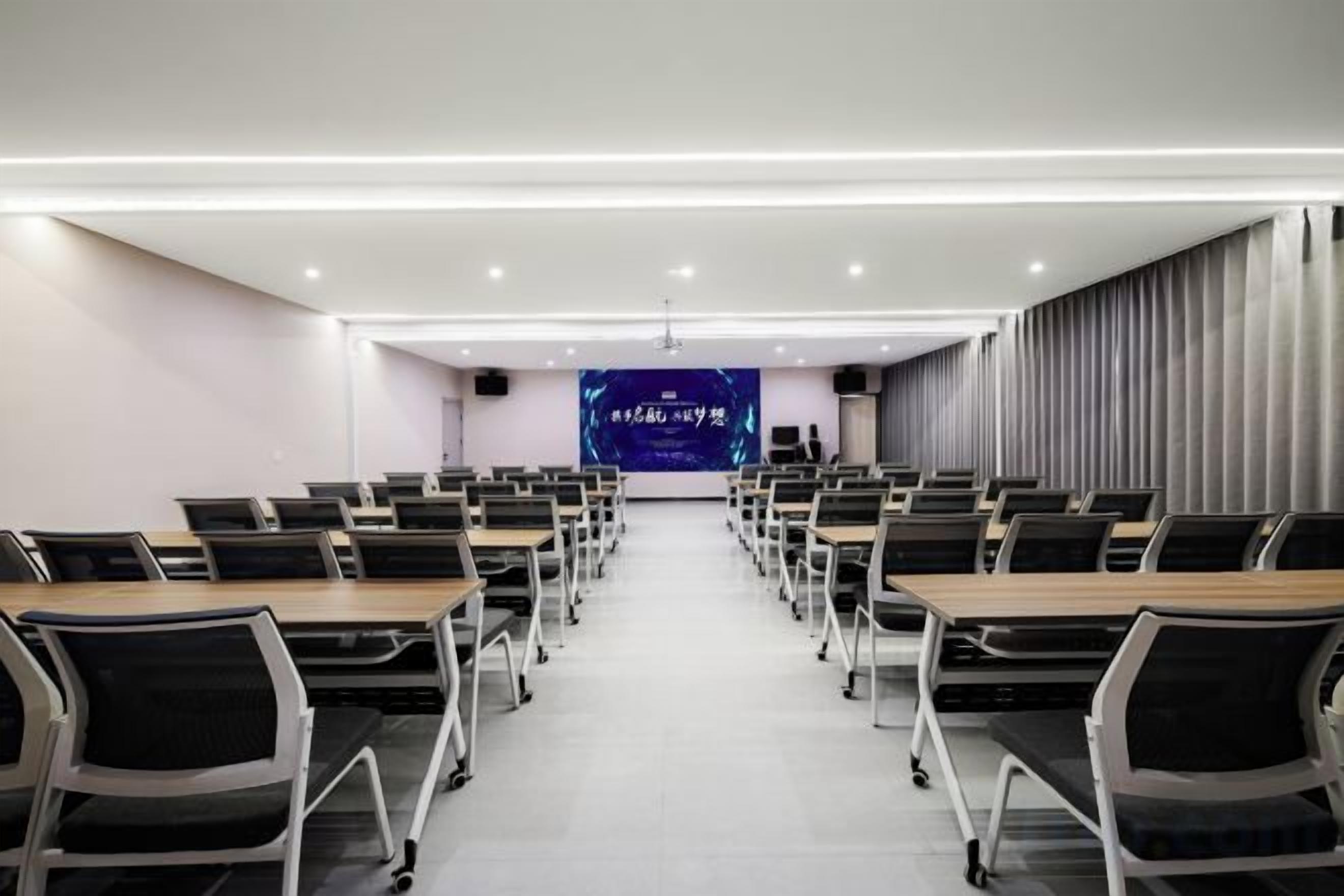 Sala de conferência com mesas e cadeiras dispostas em fileiras, uma tela para apresentação e esquema de cores neutras.