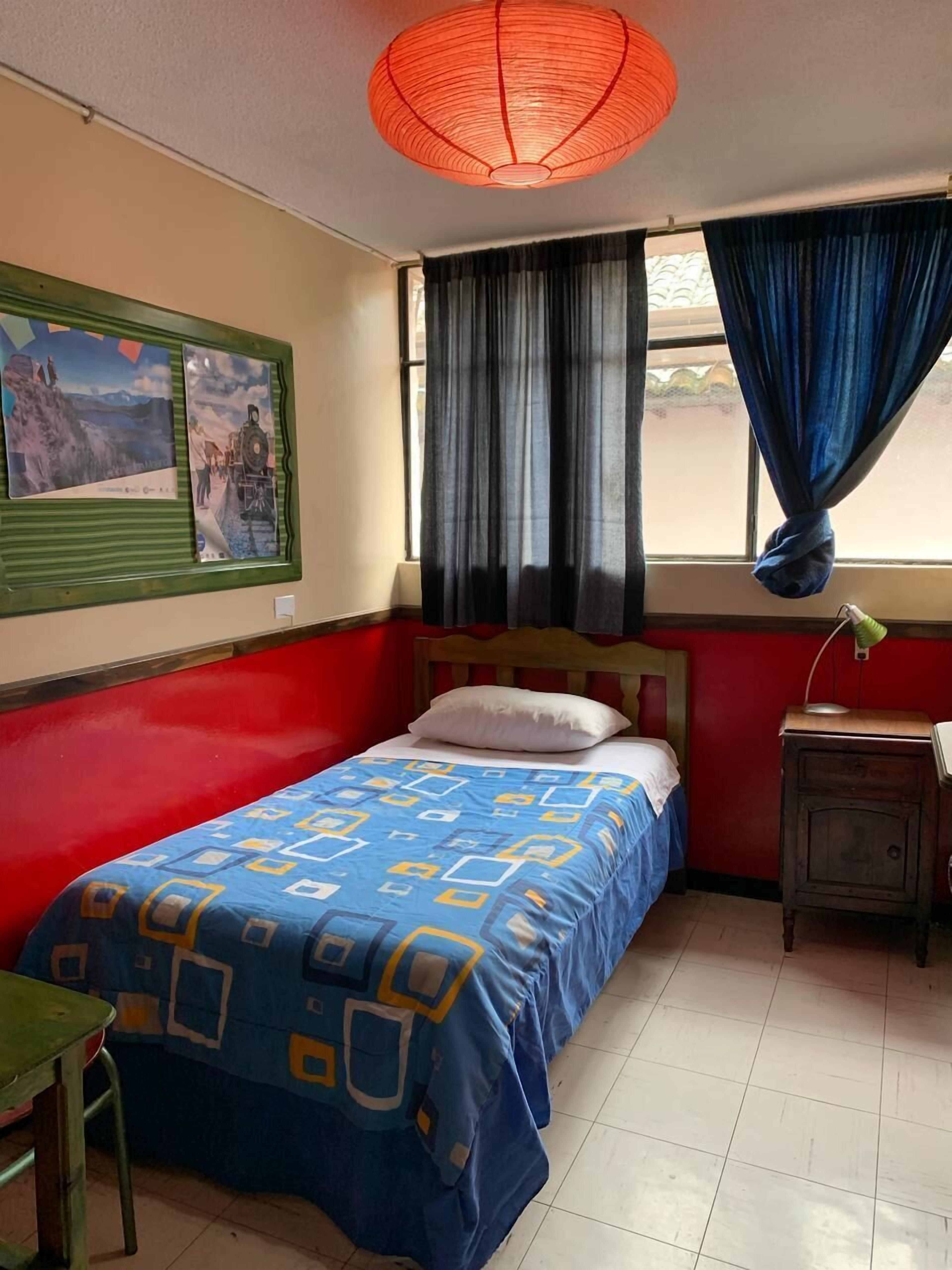 Quarto individual com uma parede vermelha, colcha de cama azul estampada, cabeceira de madeira, mesa de cabeceira com abajur e uma janela com cortinas azuis.