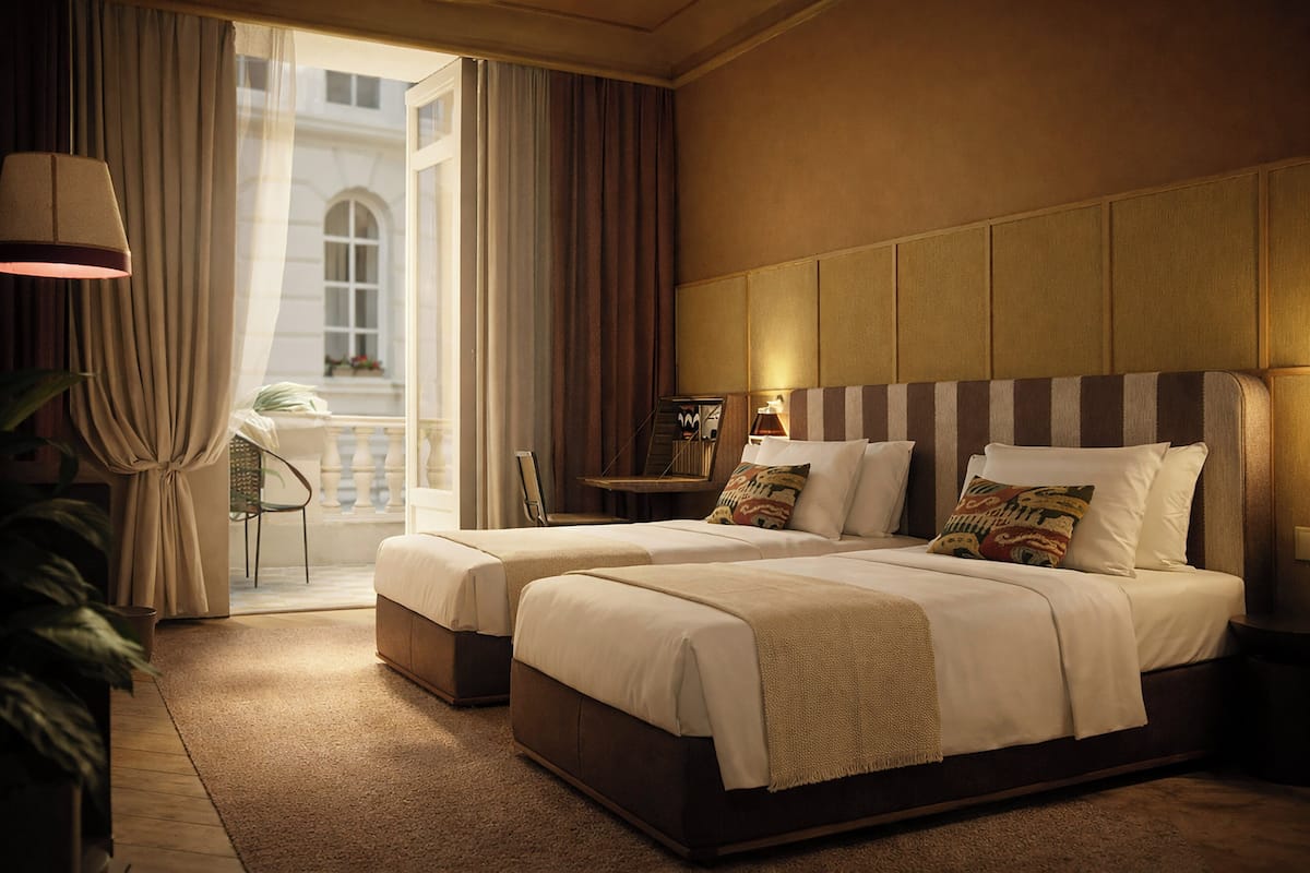 Quarto de hotel com duas camas, uma mesinha de cabeceira, um abajur, uma cadeira e vista para um edifício pela janela.
