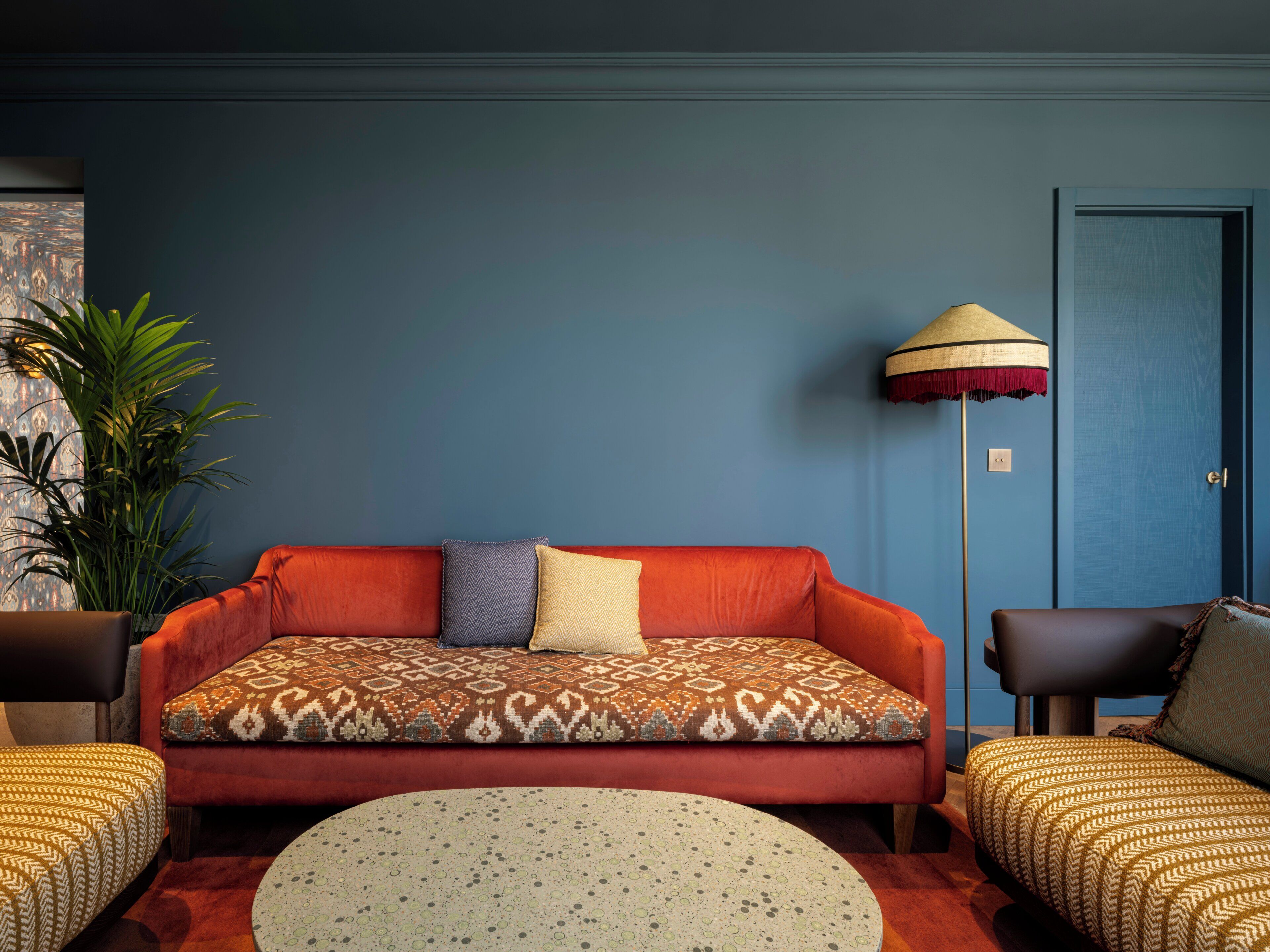Um quarto com parede azul, sofá vermelho com almofadas estampadas, mesa de centro redonda e um abajur.