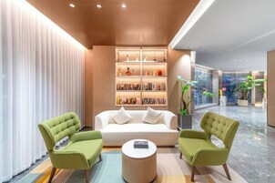 Um lobby moderno com sofá branco, poltronas verdes, mesa de centro redonda e uma estante com diversos objetos.