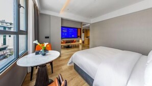 Quarto de hotel com uma cama grande, uma mesa de jantar com cadeiras, uma televisão e vista para a cidade.