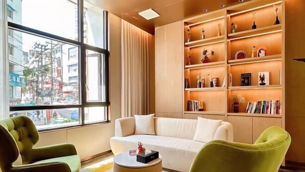 Uma sala de estar moderna com um sofá branco, duas poltronas verdes e uma mesa de centro redonda. Há uma janela grande com vista para o horizonte da cidade. Uma estante com diversos objetos está encostada na parede.