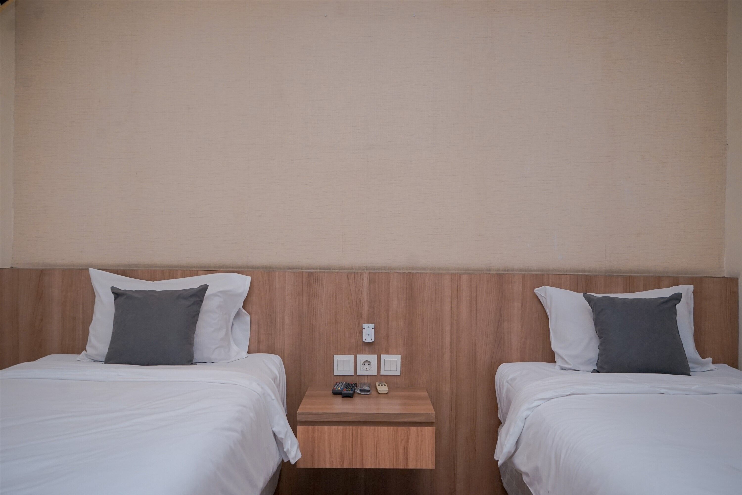 Quarto de hotel com duas camas, cada uma com roupa de cama branca e almofadas cinzas, uma cabeceira de madeira, uma pequena mesa de cabeceira com telefone e controles remotos, e uma parede lisa.