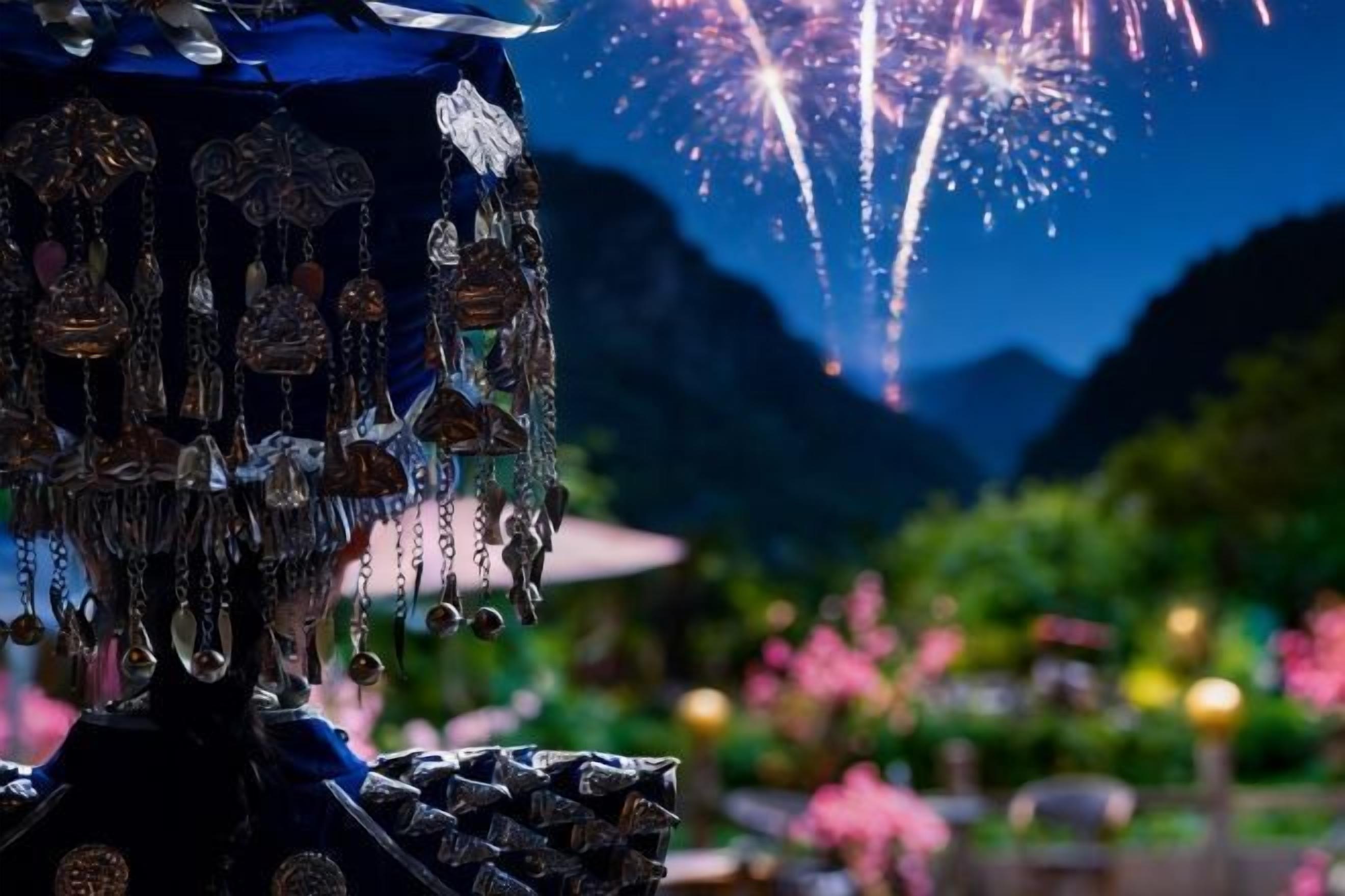Uma pessoa vestida com traje tradicional e adorno na cabeça, com fogos de artifício e uma paisagem montanhosa ao fundo.