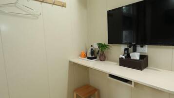 Quarto pequeno e limpo com televisão, chaleira, xícara, planta e um banquinho.