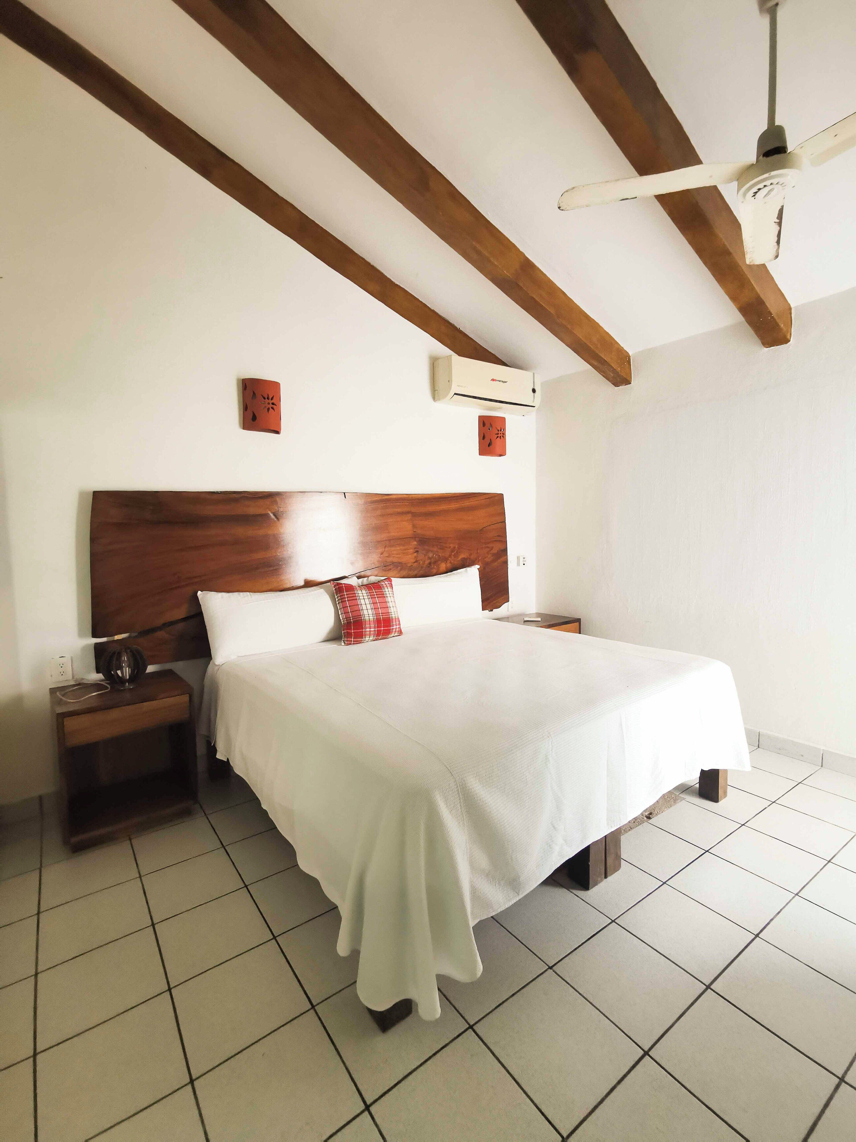 Quarto com cabeceira de madeira, colcha branca, almofada vermelha e branca, mesa de cabeceira, ar condicionado fixado na parede e ventoinha de teto.