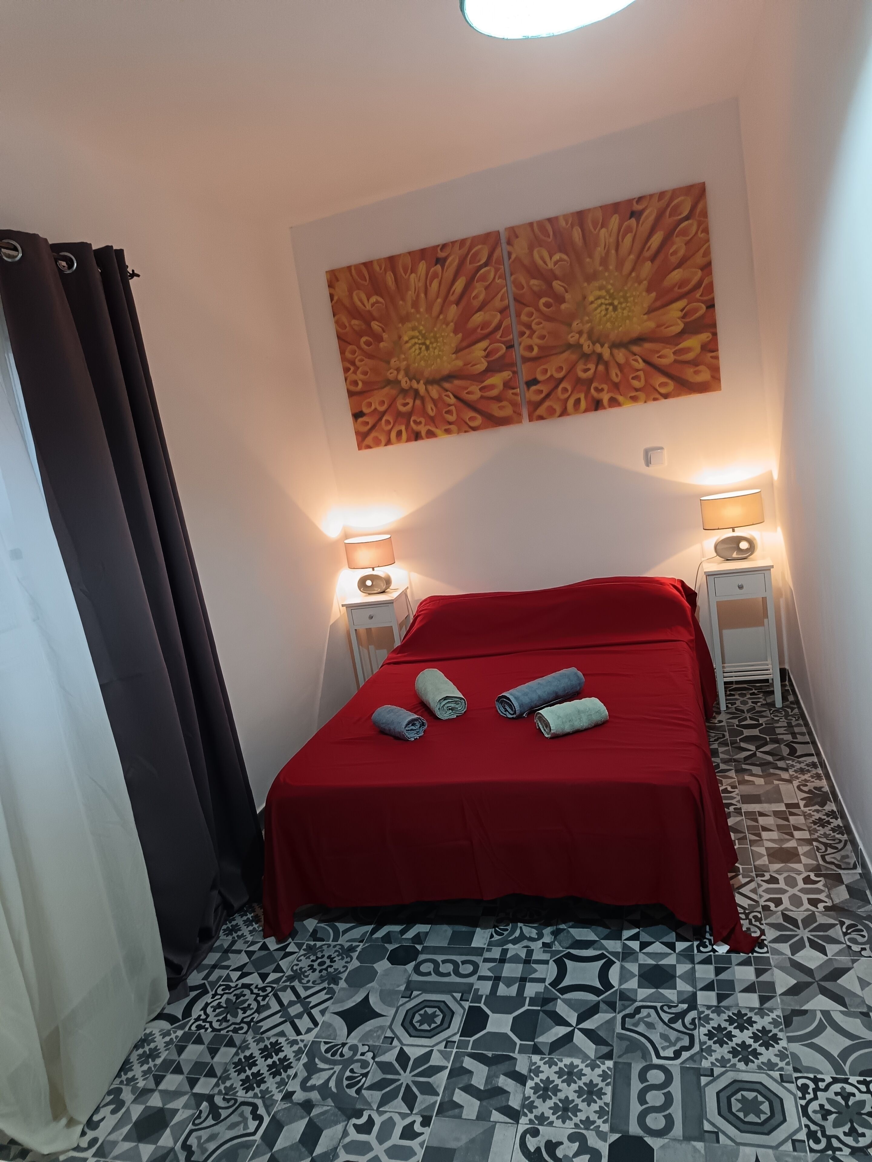 Quarto de hotel com colcha vermelha, duas luminárias de cabeceira, um quadro de flores grande na parede e ladrilhos de chão estampados.