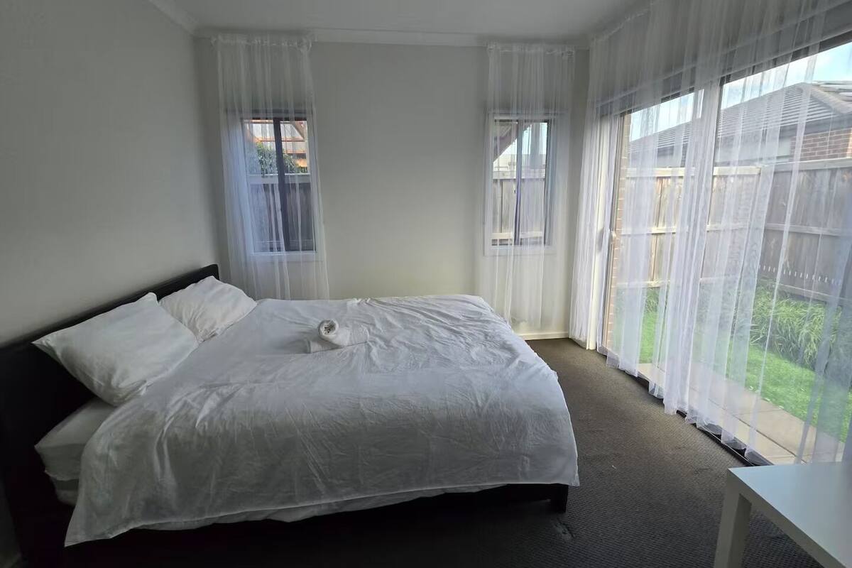Quarto com cama, duas janelas com cortinas leves e vista para um quintal, além de uma pequena mesa.
