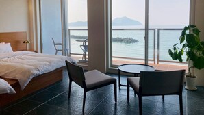 Quarto de hotel com uma cama, duas cadeiras, uma mesinha e um vaso de planta. O quarto tem janelas grandes com vista para o mar e uma ilha ao longe.