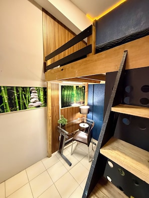 Quarto com beliche, uma pequena mesa com abajur, uma cadeira e televisão na parede.