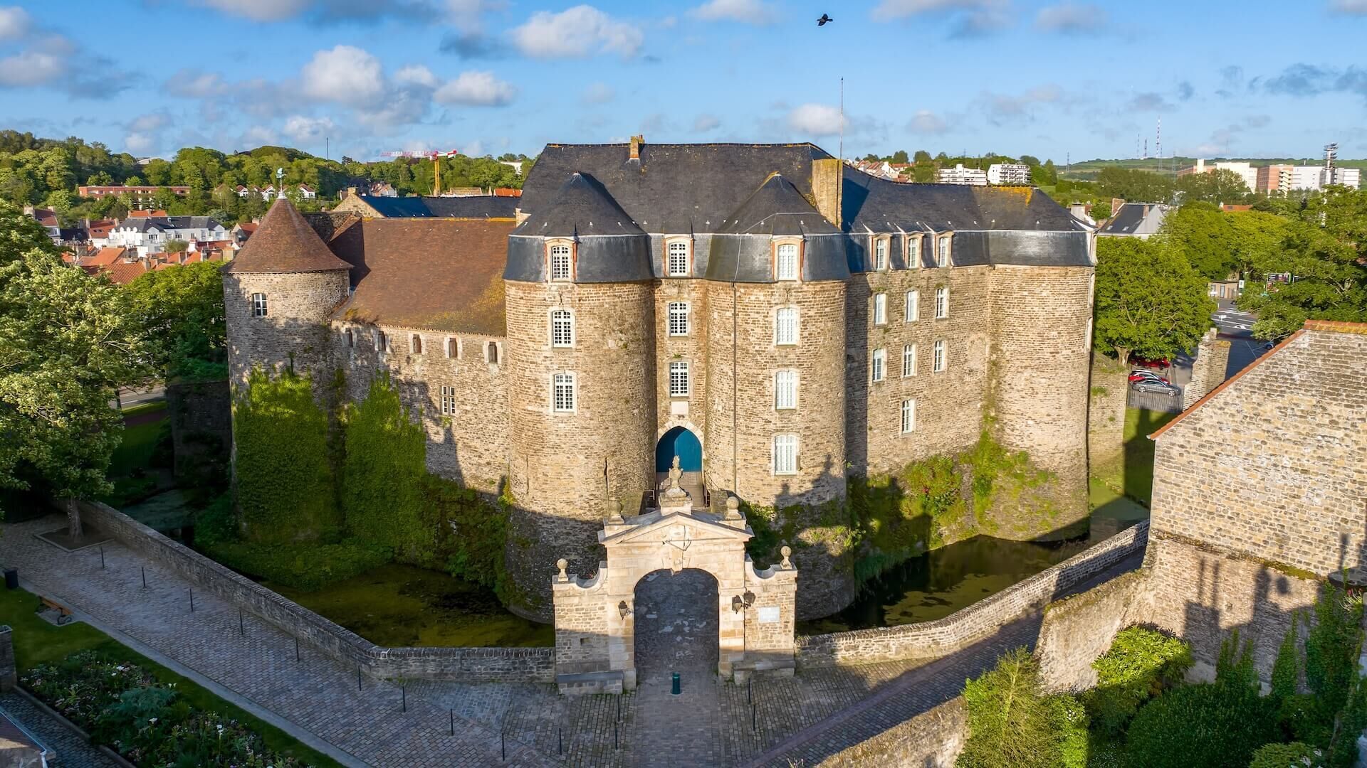 Um castelo de pedra histórico com um pátio central, rodeado por vegetação exuberante e um fosso.