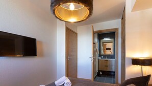 Quarto de hotel com cama, televisão de tela plana na parede, banheiro visível por uma porta aberta e abajur na mesinha de cabeceira.