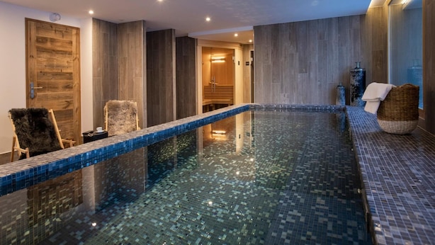 Piscina interior moderna com fundo de vidro transparente, paredes de madeira e uma área de estar com uma cadeira e um cesto.