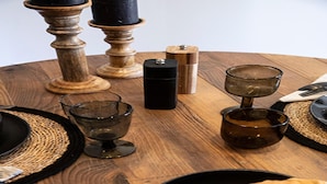 Uma mesa de madeira redonda com um prato preto, um copo, uma vela e um copo de água.