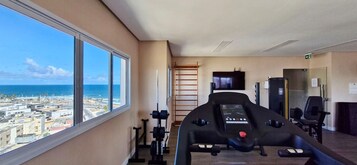 Ginásio com esteira, banco de musculação e halteres. Uma janela oferece vista para o oceano e para a paisagem urbana.
