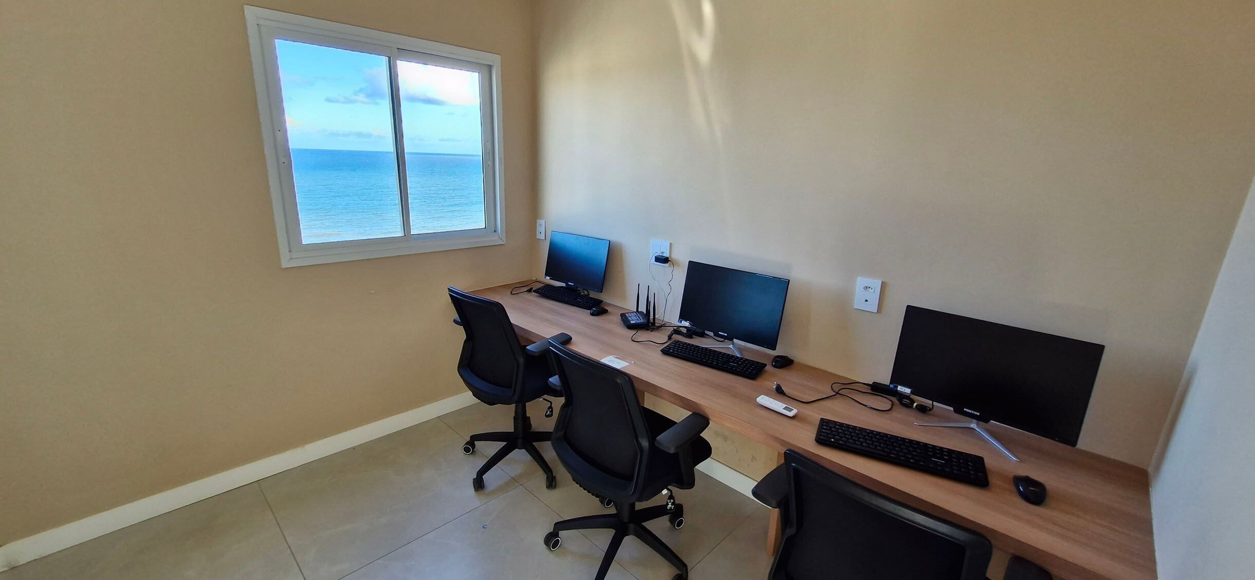 Quarto com uma grande janela com vista para o mar, uma escrivaninha de madeira com três monitores de computador, duas cadeiras de escritório pretas e um teclado na escrivaninha.