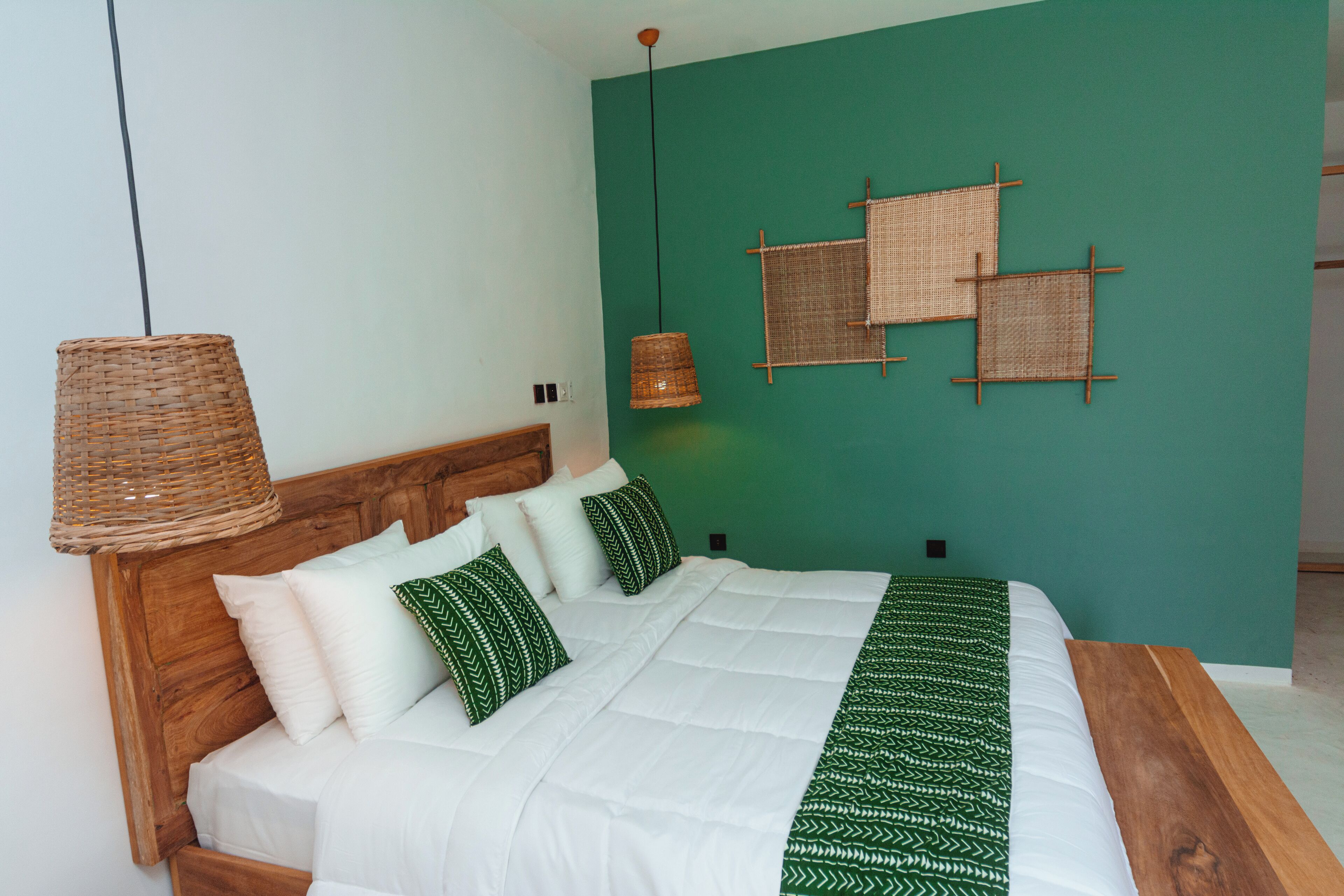Cama com roupa de cama branca e almofadas estampadas em verde, cabeceira de madeira e luminária pendente trançada.