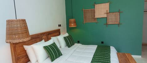 Cama com roupa de cama branca e almofadas estampadas em verde, cabeceira de madeira e luminária pendente trançada.