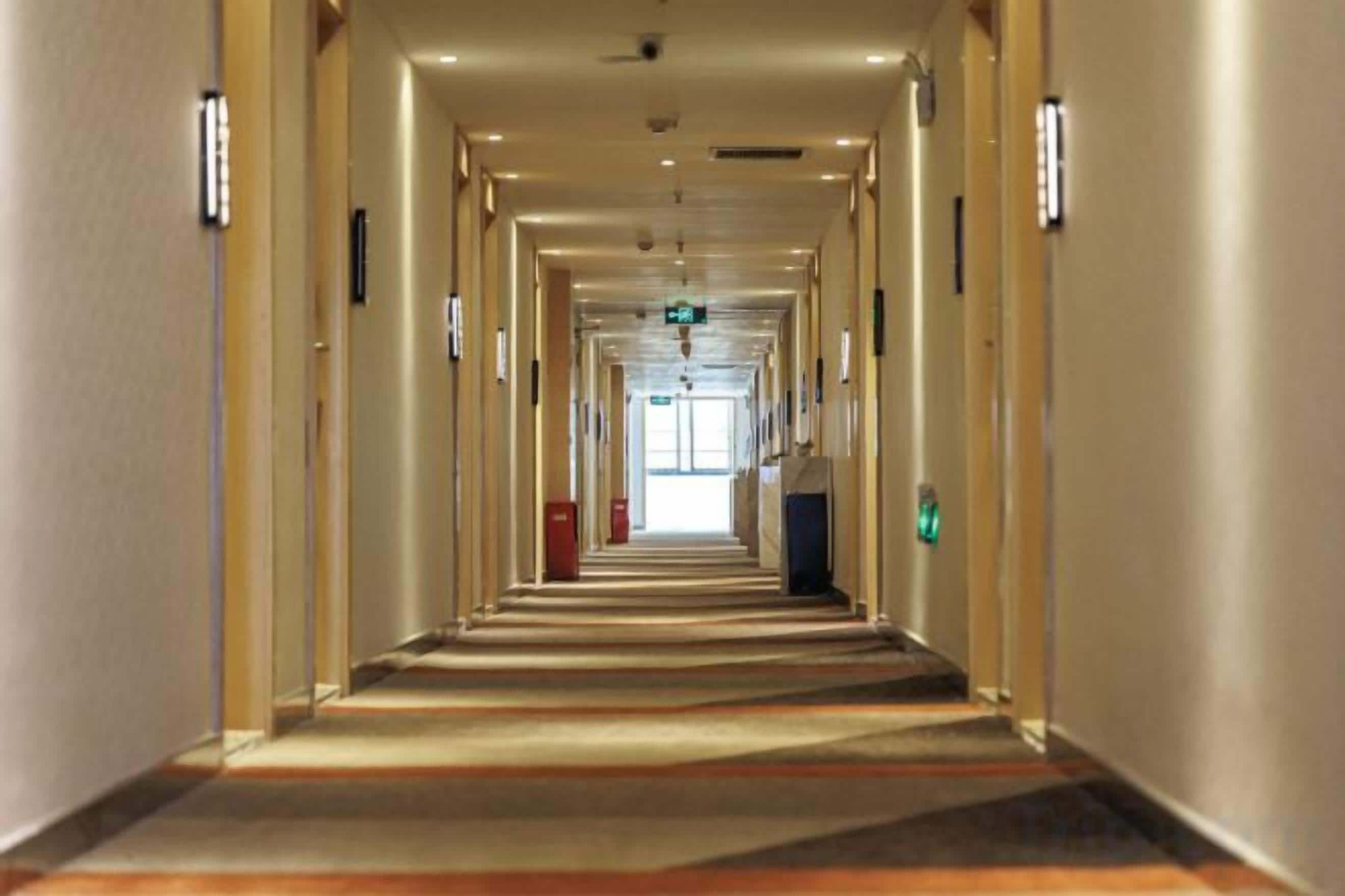 Corredor de hotel com portas em ambos os lados, piso carpetado e teto com iluminação.