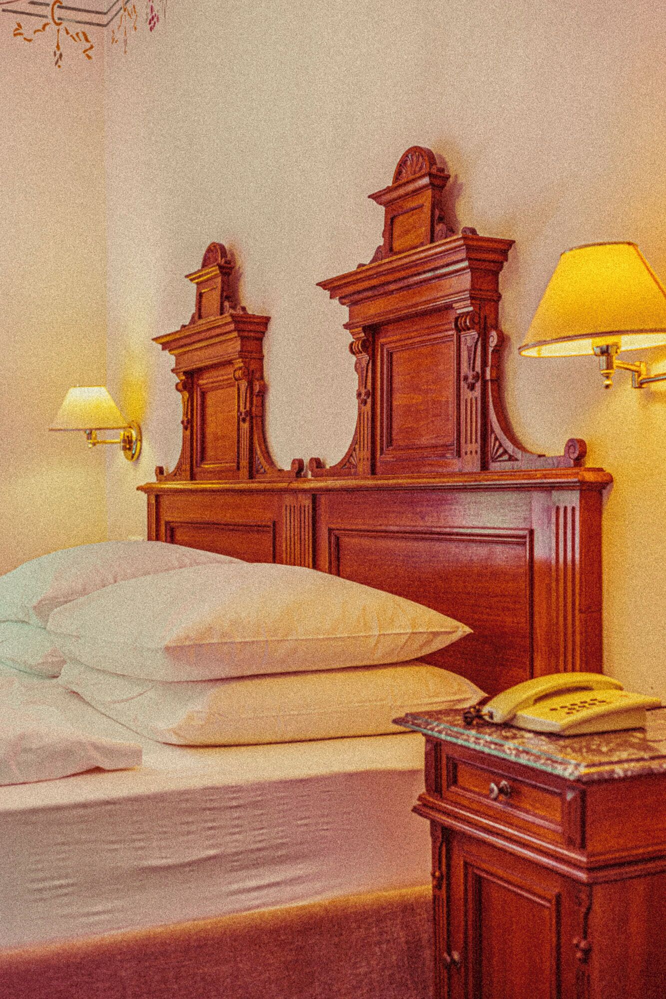 Quarto com cabeceira de madeira, duas luminárias de apoio, mesa de cabeceira com telefone e cama com almofadas.