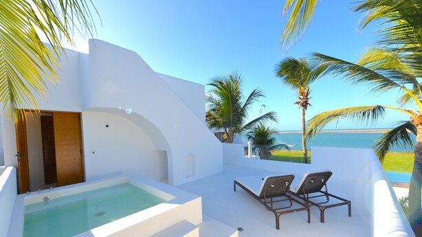Uma área de piscina moderna ao ar livre, com vista clara para o mar, duas espreguiçadeiras e uma porta de madeira que dá para um espaço coberto.