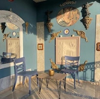 Quarto com parede azul, mesa de madeira, duas cadeiras, espelho decorativo e esculturas na parede.