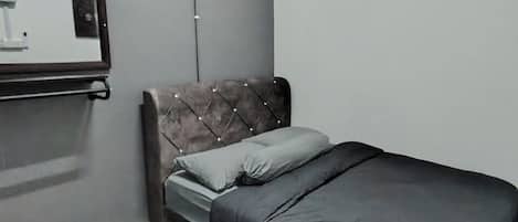 Quarto com cama, ar condicionado fixado na parede e uma pequena prateleira com um quadro emoldurado.