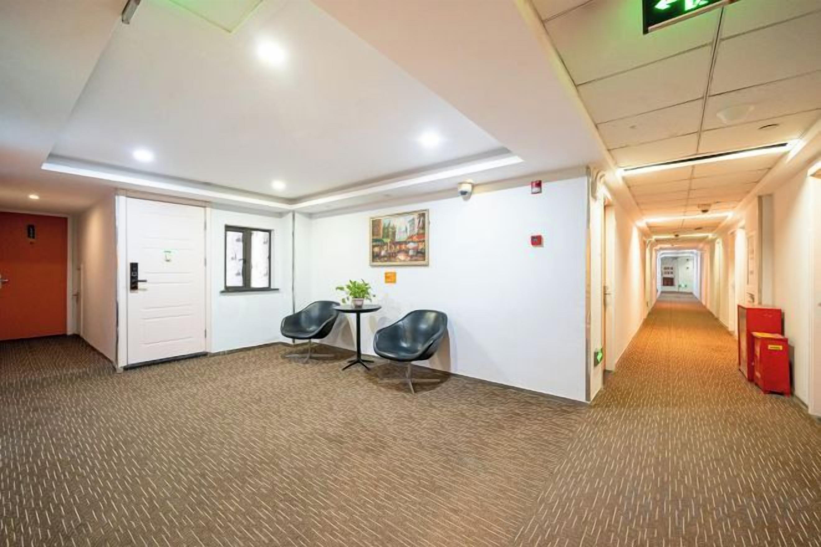 Um saguão de hotel com piso carpete, uma mesa de recepção, duas cadeiras e um quadro na parede.