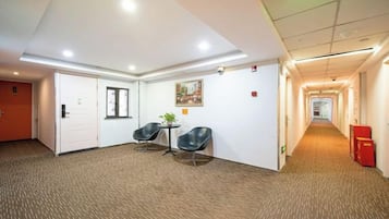 Um saguão de hotel com piso carpete, uma mesa de recepção, duas cadeiras e um quadro na parede.