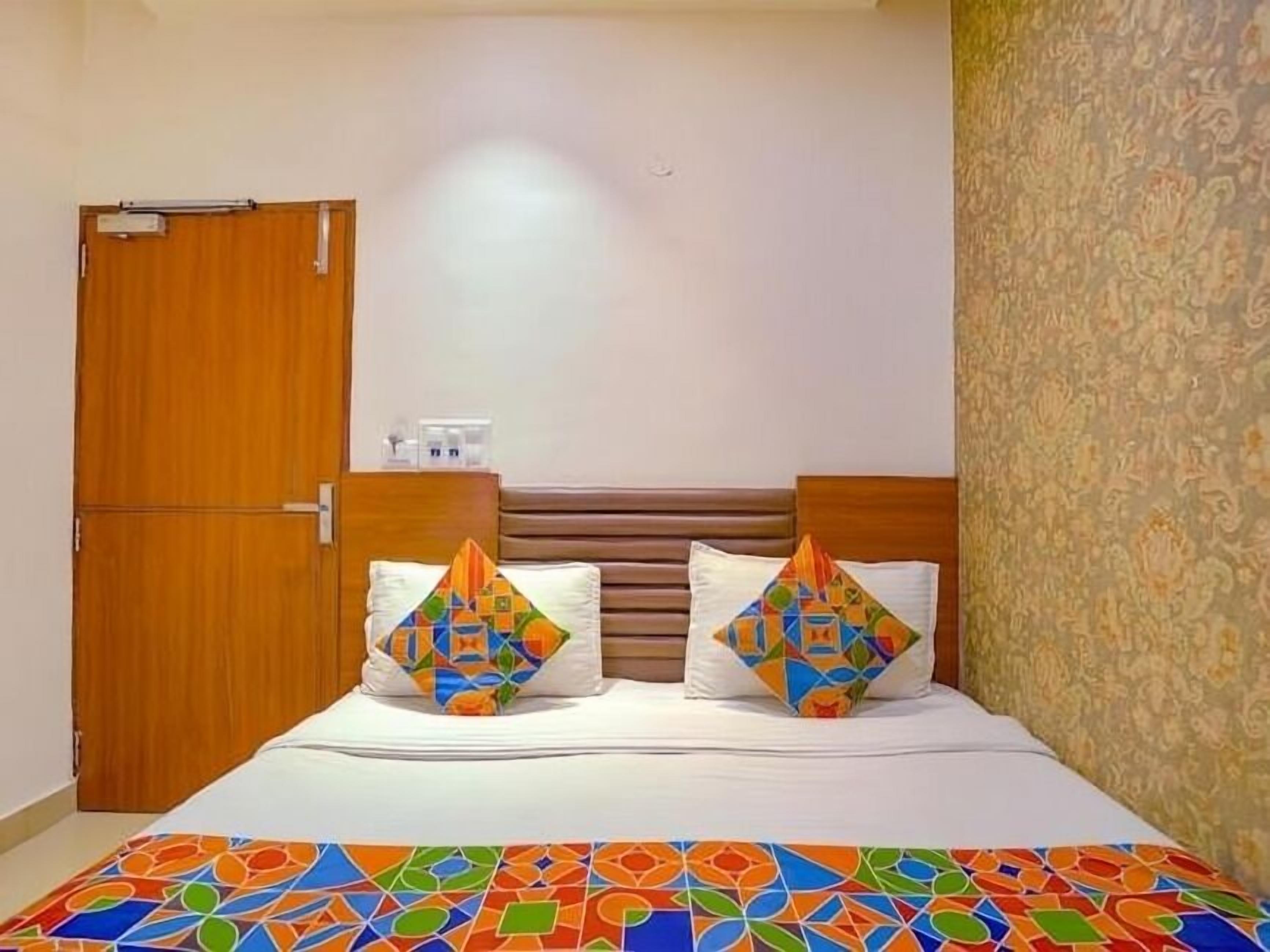 Quarto de hotel com uma cama, duas almofadas com padrões geométricos, uma porta de madeira e uma parede estampada.