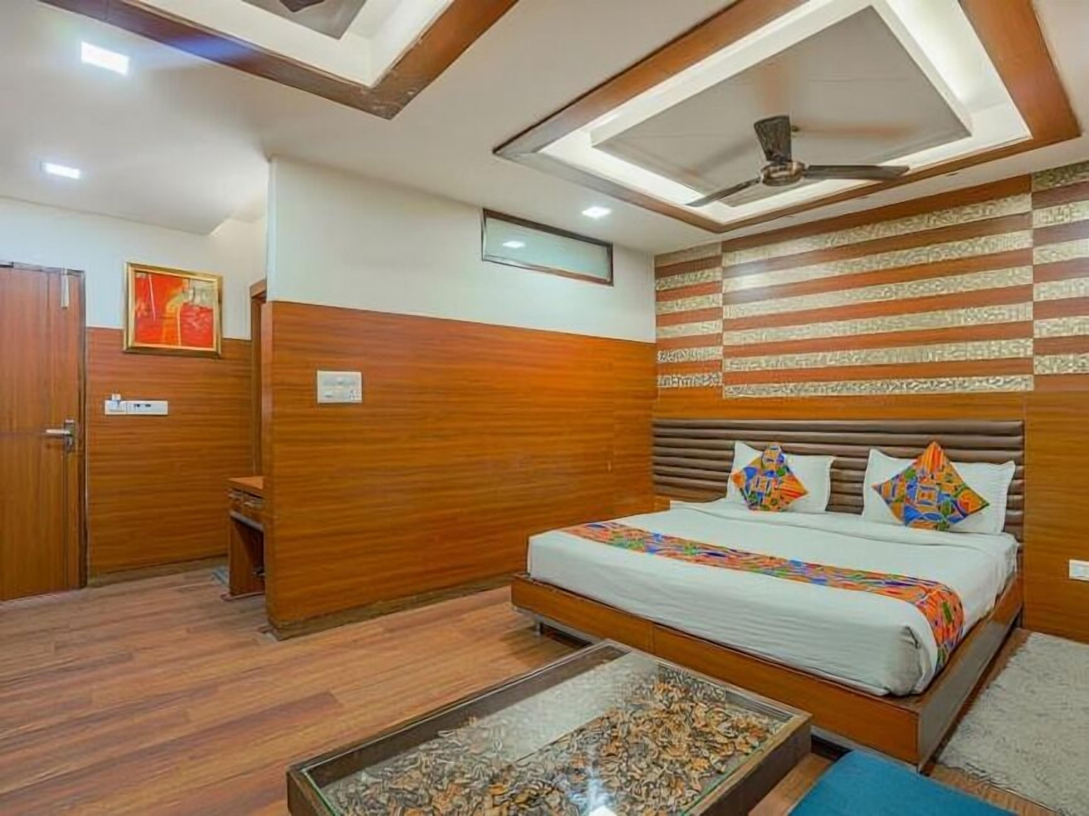 Quarto de hotel com chão de madeira, cama com almofadas coloridas, ventilador de teto e uma pequena mesa com um objeto decorativo.