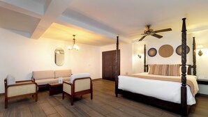 Quarto de hotel com cama de dossel, sofá, duas poltronas, ventoinha de teto e luminárias fixadas na parede.