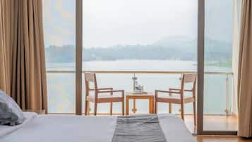 Quarto de hotel com uma cama grande, duas cadeiras, uma mesinha e vista para um lago através de grandes janelas.