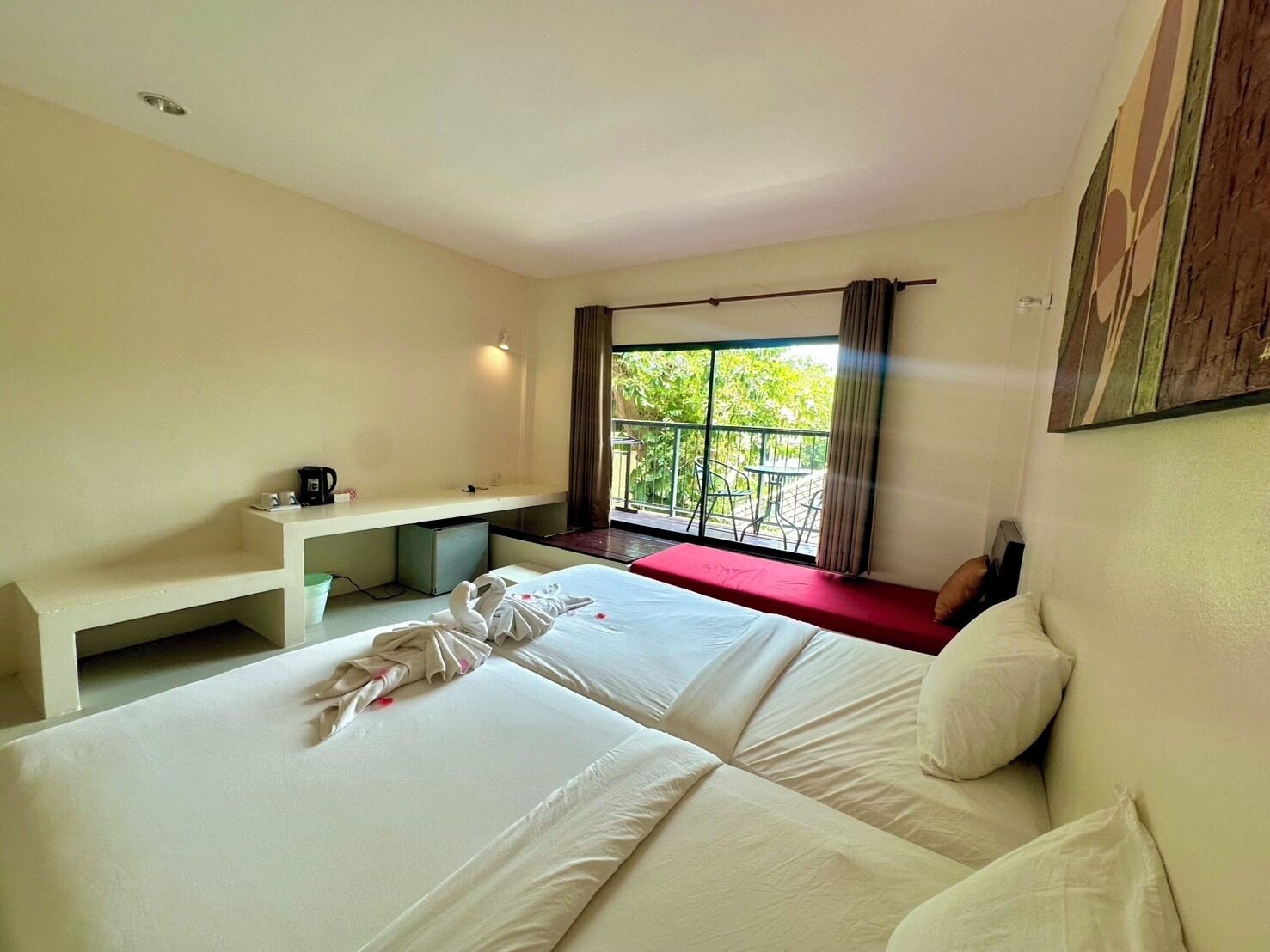 Quarto de hotel com duas camas, uma escrivaninha, uma cadeira, varanda com vista e um quadro na parede.
