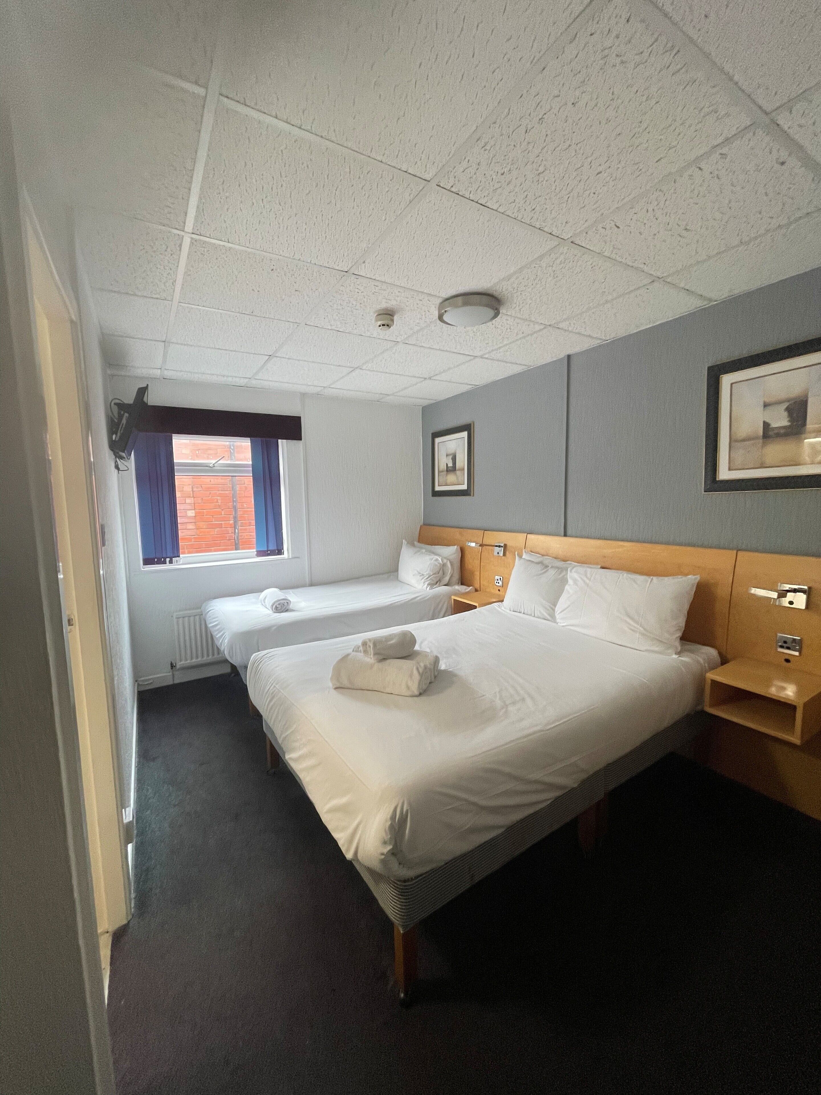 Quarto de hotel com duas camas de solteiro, cabeceira de madeira, mesa de cabeceira, quadro pendurado na parede e uma janela com cortinas.