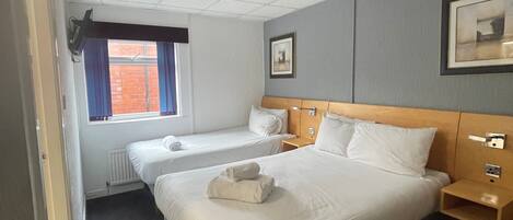 Quarto de hotel com duas camas de solteiro, cabeceira de madeira, mesa de cabeceira, quadro pendurado na parede e uma janela com cortinas.