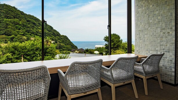 Uma sala de jantar com cadeiras de vime e uma mesa de madeira, com vista para uma encosta verdejante e o mar.