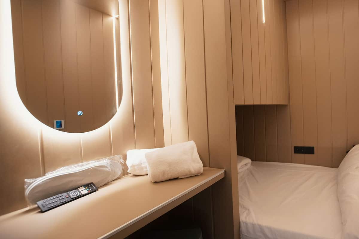 Quarto de hotel moderno com parede revestida em madeira, cama com roupa de cama branca, mesa de cabeceira com controle remoto e espelho embutido.