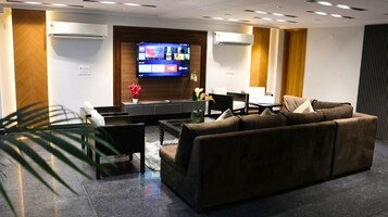 Um lobby moderno com um sofá, uma mesa de centro e uma TV na parede exibindo diversos canais.
