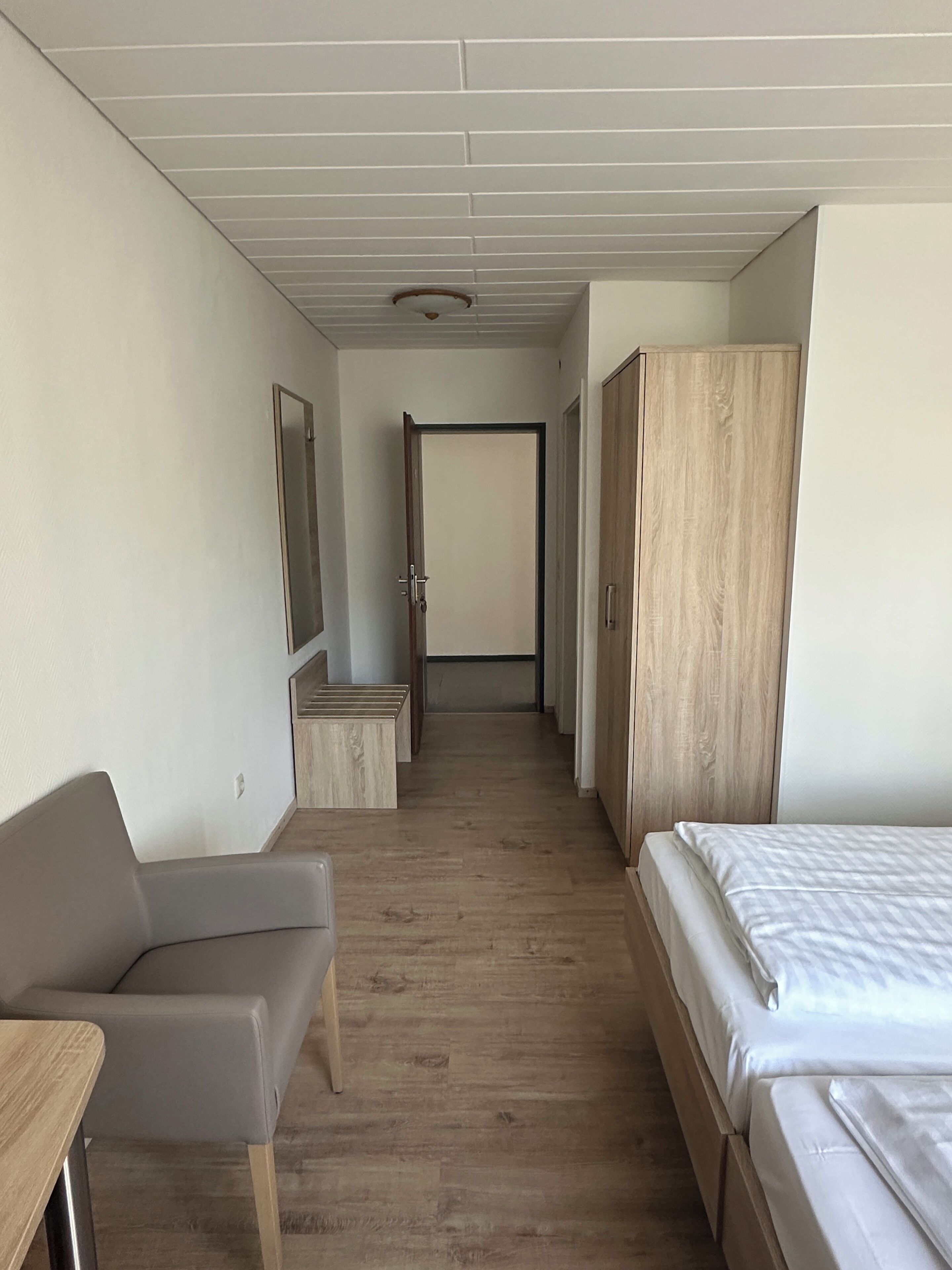 Quarto de hotel com cama, escrivaninha, cadeira, guarda-roupa e porta que dá para outro espaço.