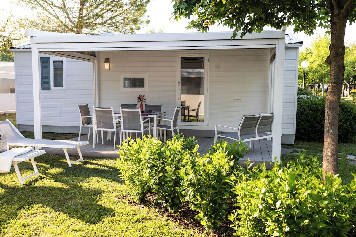 Uma casa móvel branca com um espaço de pátio com mesa e cadeiras, cercada por vegetação.