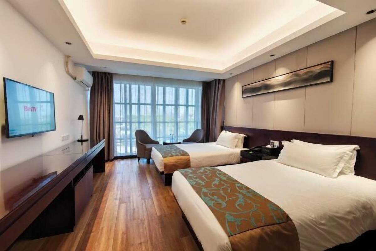 Quarto de hotel com duas camas, uma televisão, uma escrivaninha e uma janela grande.