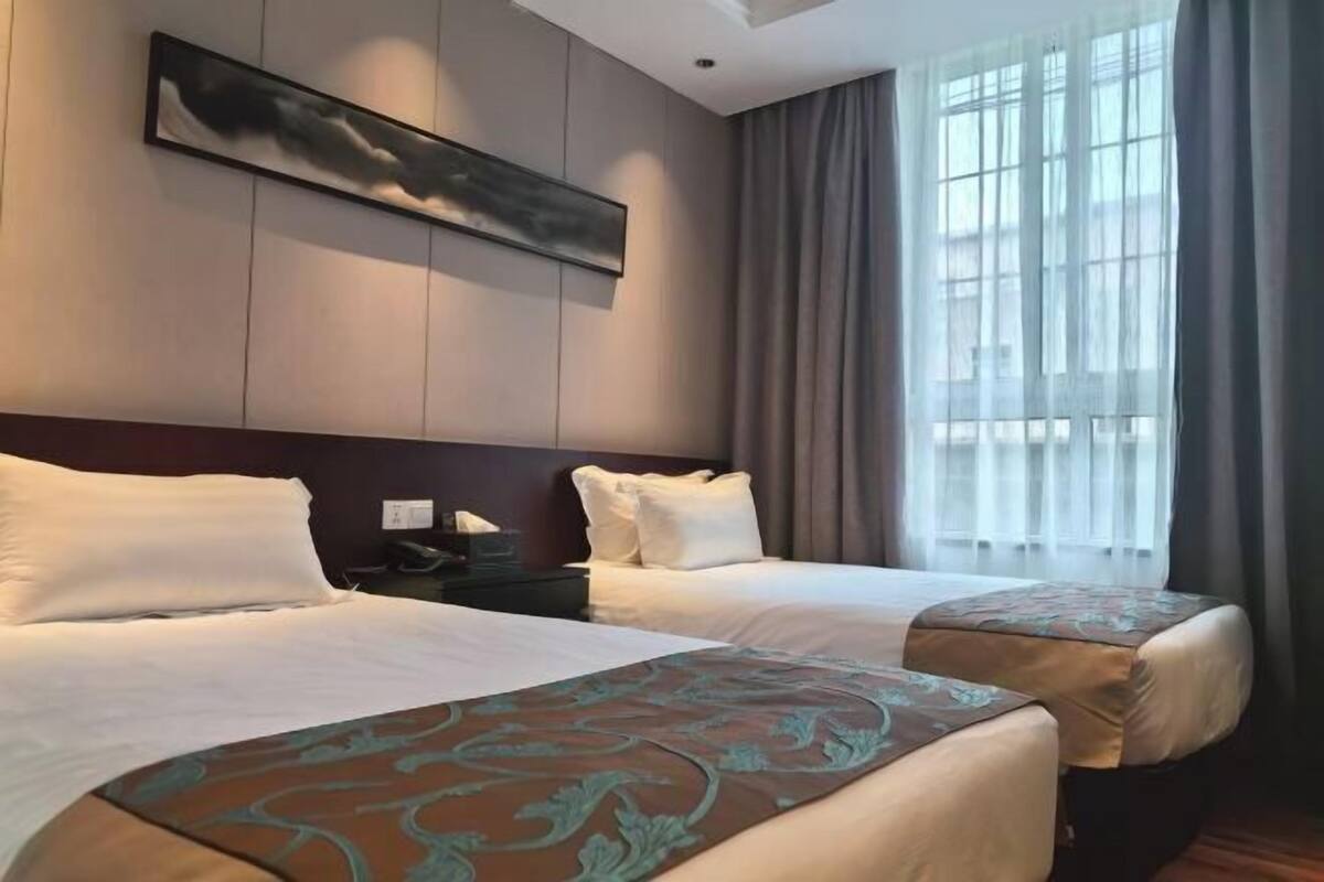 Quarto de hotel com duas camas, uma mesa de cabeceira, um quadro na parede e uma janela com cortinas.
