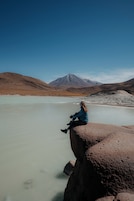 Uma pessoa sentada em uma rocha à beira de um lago calmo, com montanhas ao fundo.
