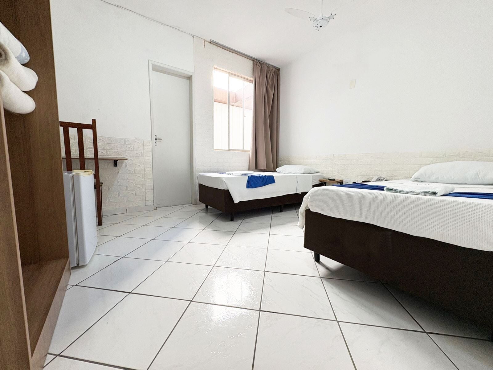 Quarto de hotel com duas camas, uma janela com cortinas, uma porta e um ventilador de teto.