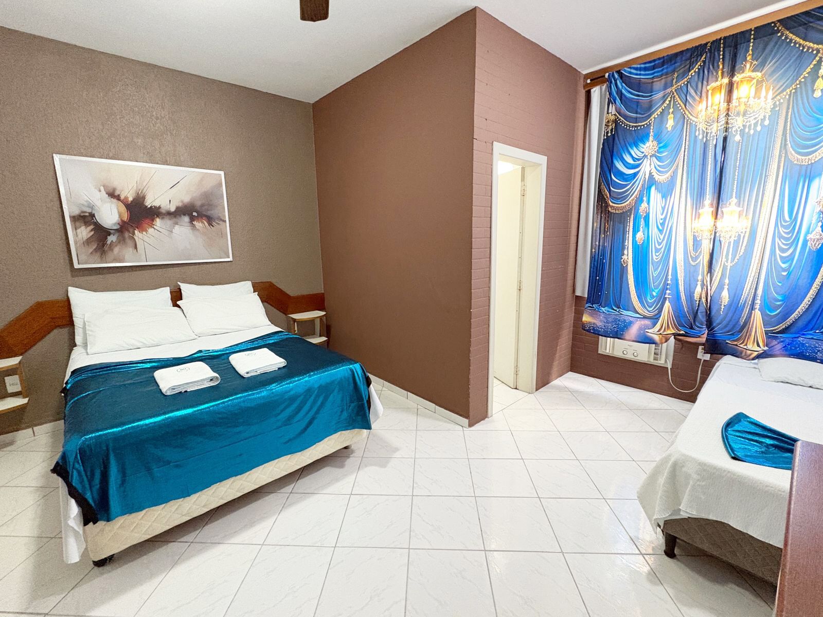 Quarto de hotel com cama, mesa de cabeceira, uma cadeira, uma janela grande com cortinas e um quadro na parede.