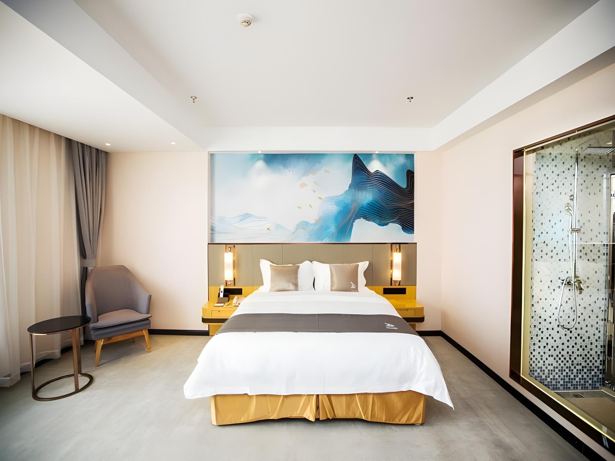 Quarto de hotel moderno com uma cama grande, uma mesinha de cabeceira, uma cadeira, um quadro na parede e box de vidro.